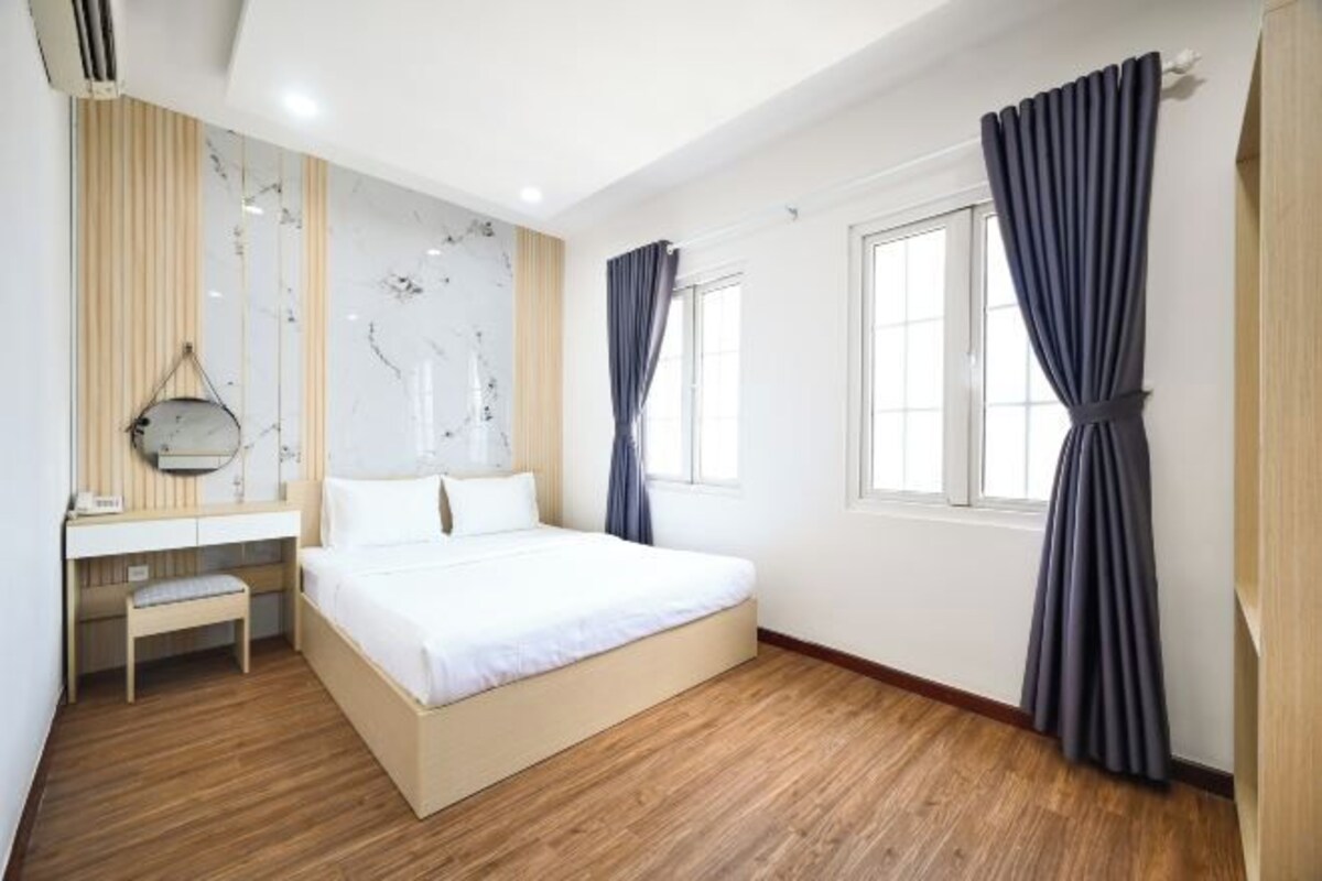 Quarto de hotel com piso de madeira, uma cama com roupa de cama branca, mesa de cabeceira com abajur, espelho e duas janelas com cortinas escuras.