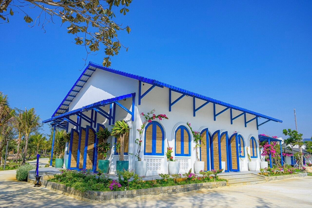 Um edifício branco com detalhes em azul e janelas em arco, rodeado por vegetação e palmeiras.