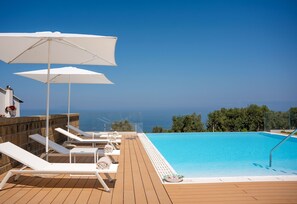 Uma área à beira da piscina com espreguiçadeiras e guarda-sóis, um deck de madeira e vista para o mar.