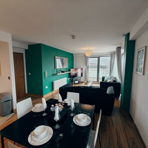 Uma sala de estar moderna com uma mesa de jantar preta para quatro pessoas, um sofá, uma TV e uma grande janela com cortinas.