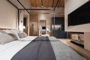 Quarto de hotel moderno com uma cama grande, televisão e área de cozinha visível através de uma porta aberta.