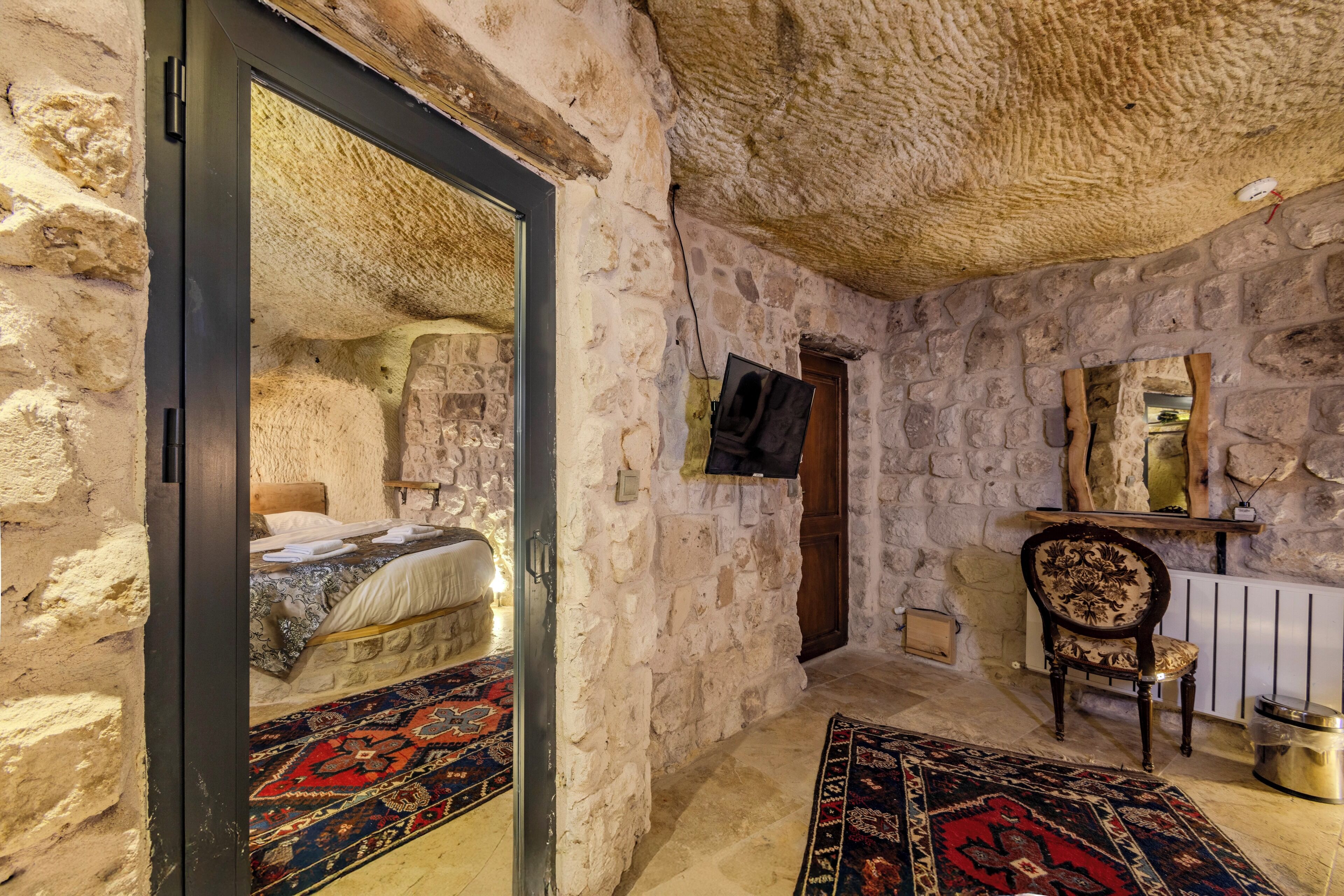 Quarto com paredes de pedra, uma cama, uma cadeira de madeira, um espelho e uma televisão.