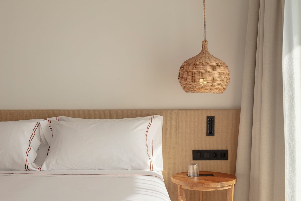 Cama bem arrumada com lençóis brancos, cabeceira de madeira e uma pequena mesa de cabeceira de madeira com um recipiente de vidro.