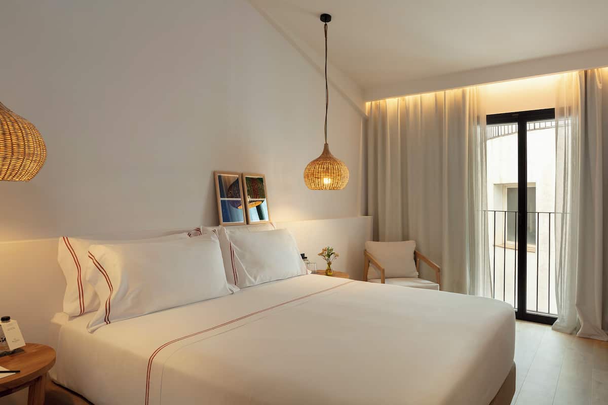 Quarto de hotel com uma cama grande, mesa de cabeceira de madeira, luminária pendente de vime, uma cadeira e vista para o exterior por uma porta deslizante.