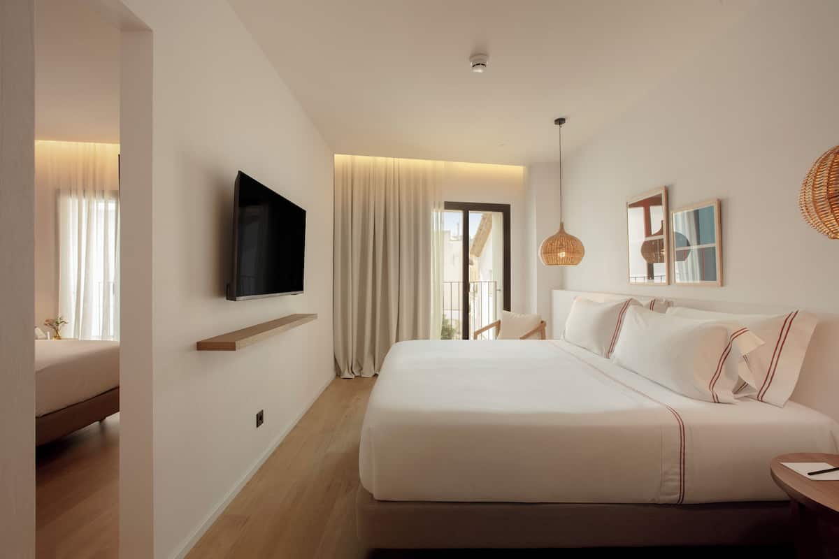 Quarto de hotel moderno com uma cama grande, televisão de tela plana na parede, mesa de cabeceira de madeira e uma janela ampla com cortinas.