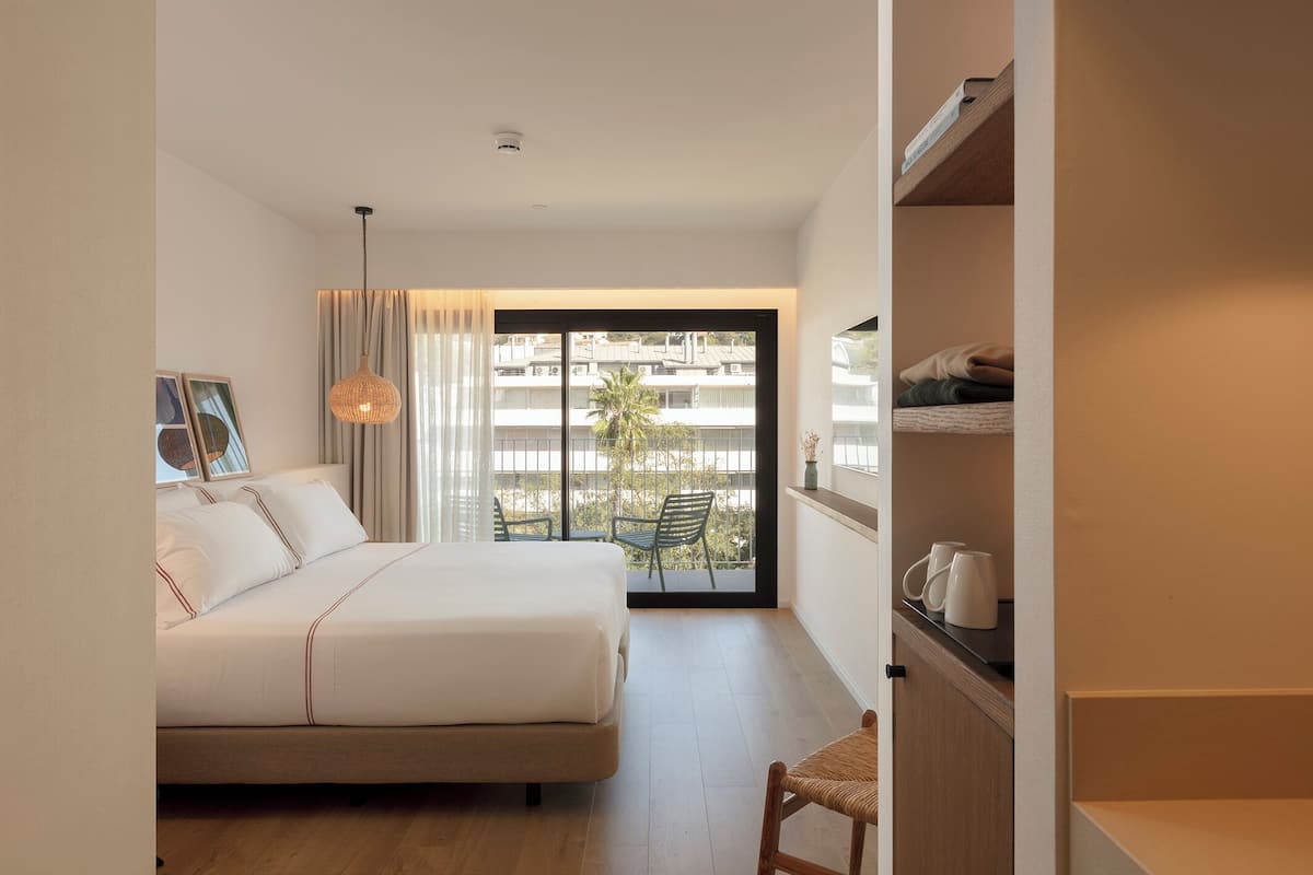 Quarto com cama, uma cadeira, prateleira com xícaras e varanda com vista para edifícios e uma palmeira.