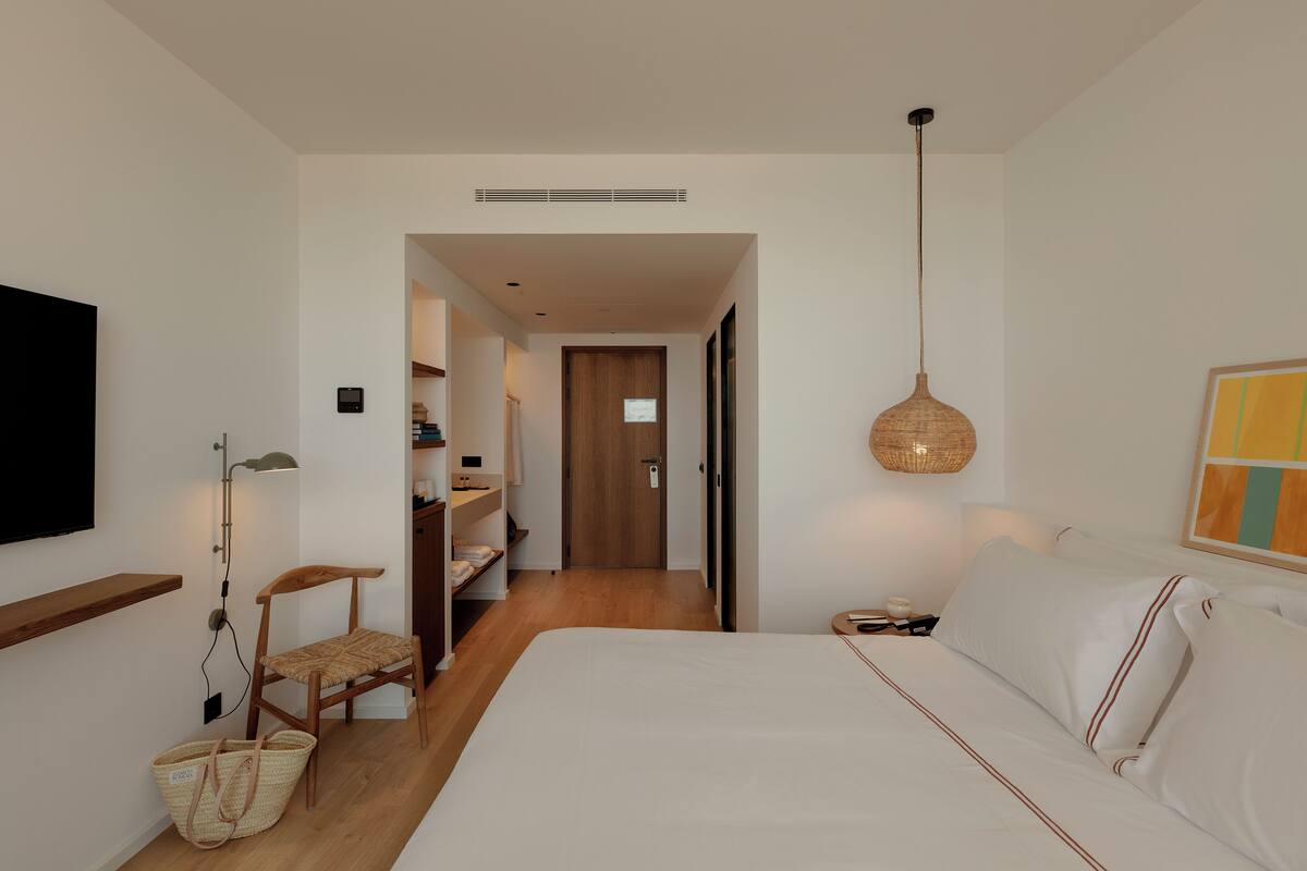 Quarto de hotel com uma cama grande, uma cadeira de madeira, uma mesa de trabalho e uma TV fixada na parede.