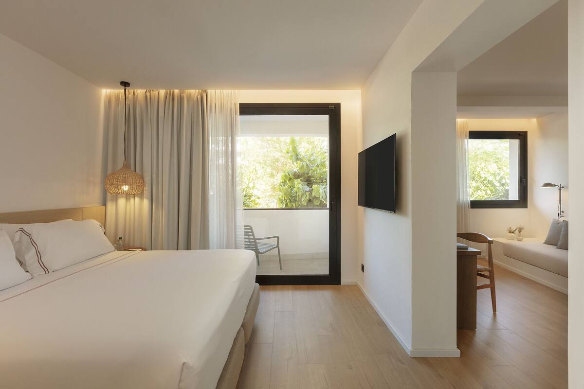 Quarto de hotel com cama, escrivaninha, cadeira, televisão e varanda com cortinas.