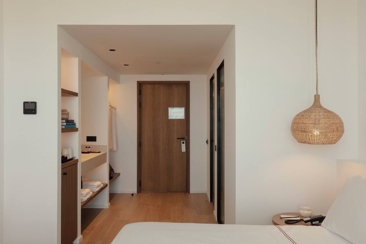 Quarto de hotel com porta de madeira, mesa de cabeceira com abajur e cama com roupa de cama branca.