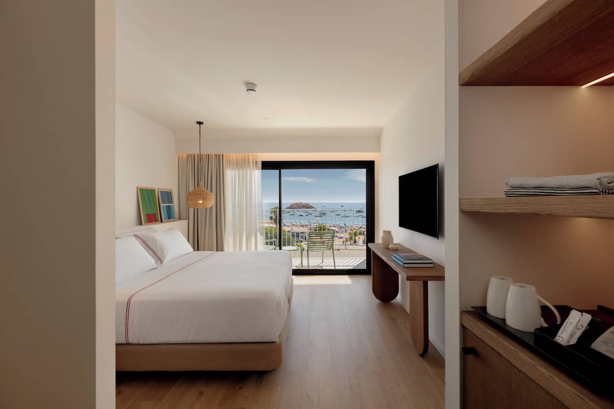Quarto de hotel moderno com uma cama grande, televisão, escrivaninha e vista da cidade por uma janela ampla.