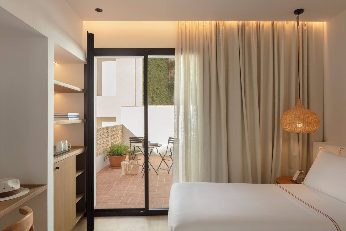 Quarto de hotel moderno com uma cama grande, área de jantar com mesa e cadeiras, pequena cozinha e porta de vidro deslizante que dá para um pátio externo com planta em vaso e cadeira de vime.