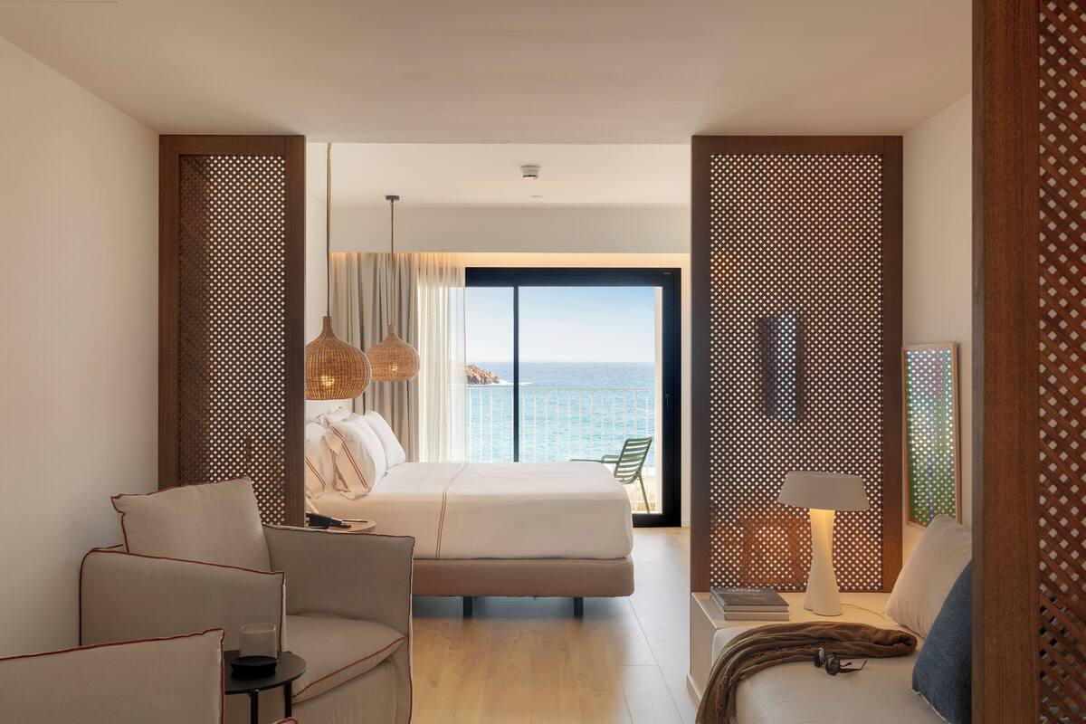 Quarto de hotel com uma cama, duas poltronas, uma mesa pequena, uma luminária e vista para o mar através de uma janela ampla.
