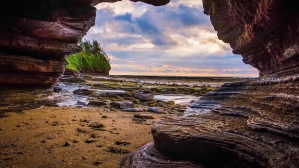 Formação rochosa natural com a entrada de uma gruta, uma praia arenosa e uma vista distante do céu.