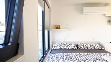 Quarto de hotel com uma cama grande, uma janela com persianas, ar condicionado fixado na parede e uma pequena estante com um porta-retrato.