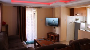 Uma sala de estar com televisão de tela plana, mesa de centro de madeira, sofá e área de cozinha com armários e geladeira.