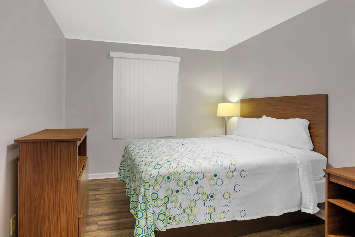 Quarto de hotel com cama, mesa de cabeceira de madeira, escrivaninha e janela com persianas.
