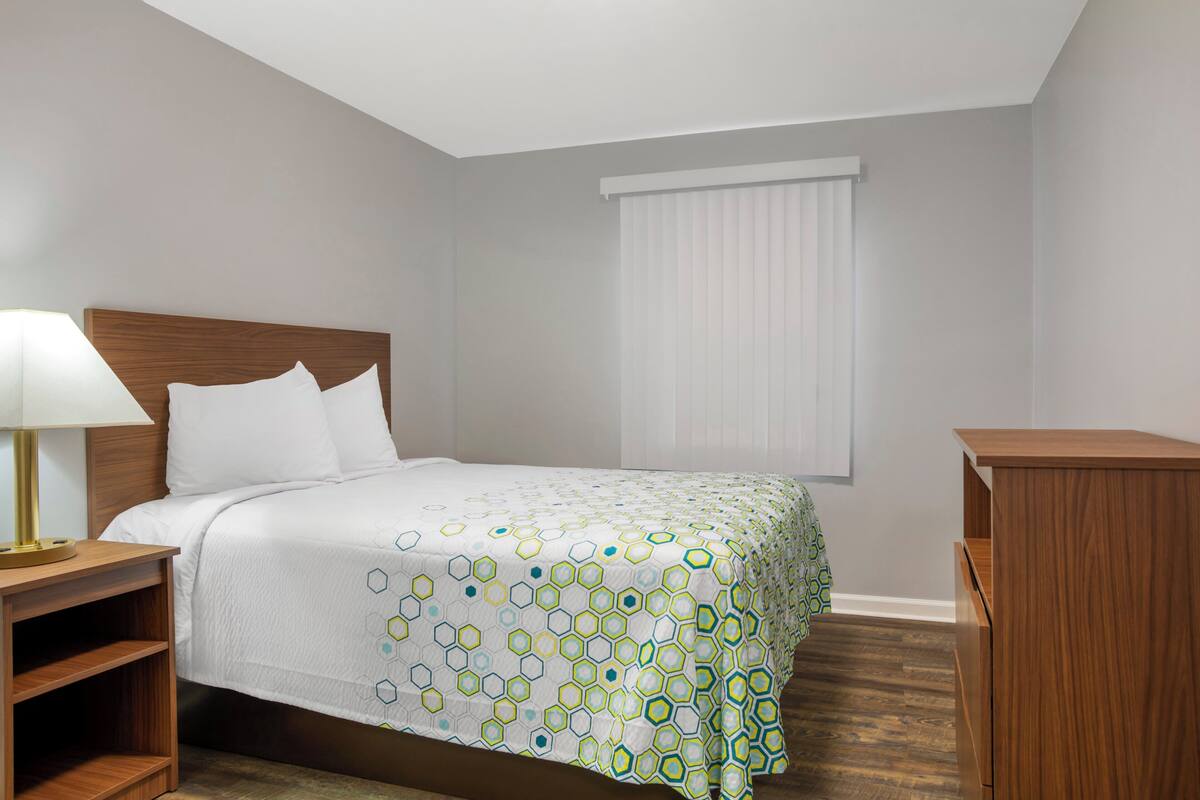 Cama bem arrumada com colcha de padrão geométrico, luminária de mesa e mesa de cabeceira de madeira.