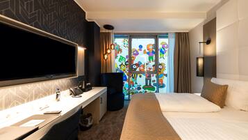 Quarto de hotel com cama, escrivaninha com telefone, televisão e janela com arte colorida.