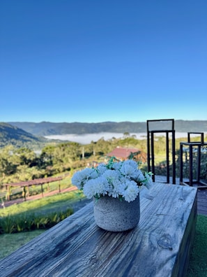 Um vaso de pedra com flores brancas sobre um banco de madeira, com vista para uma paisagem deslumbrante com montanhas e um céu azul límpido.