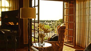 Um quarto com um piano, uma mesa com uma lâmpada e um vaso, e vista para o verde através de uma janela.