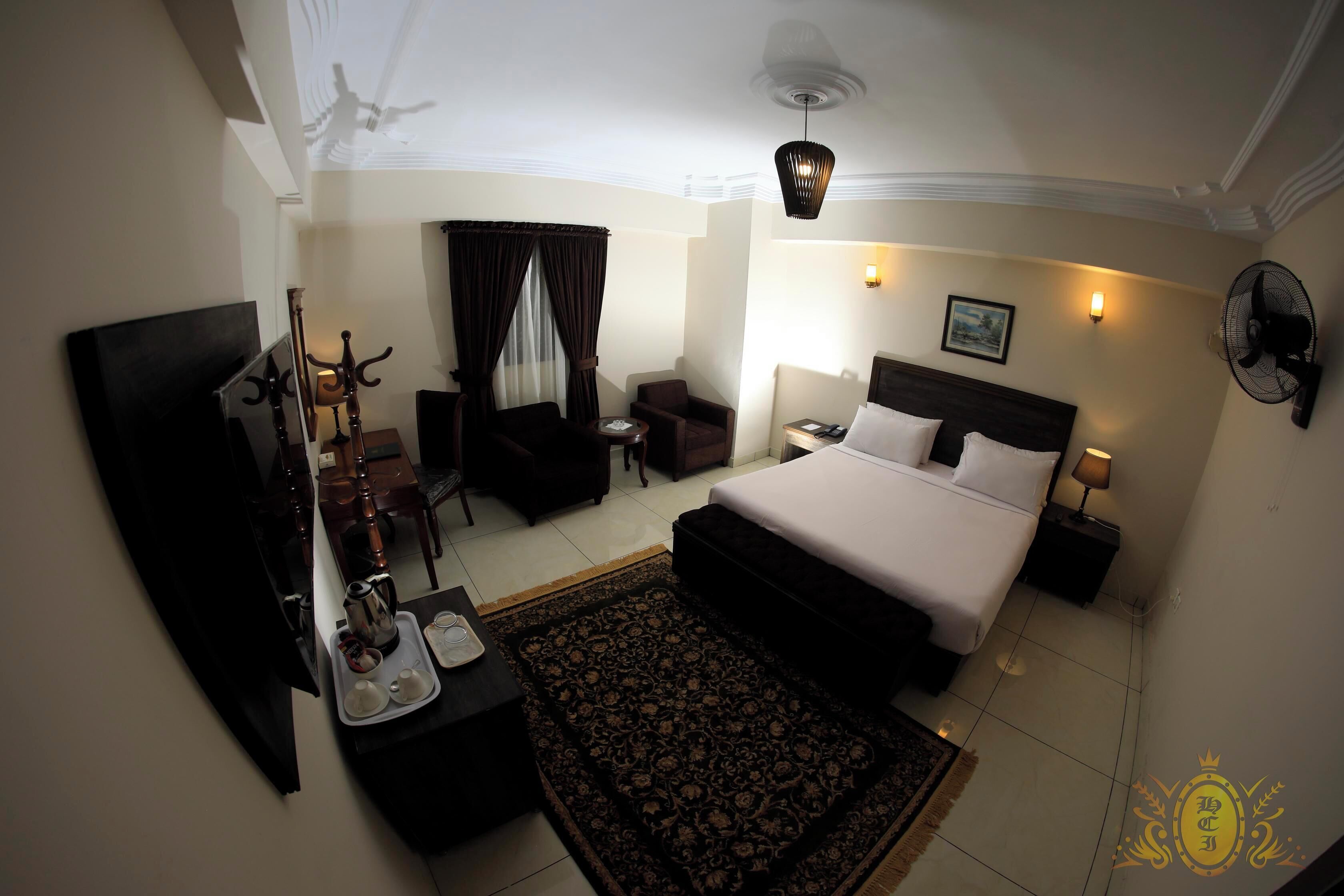 Quarto de hotel com cama, mesa de cabeceira, abajur, ventilador e um tapete decorativo.