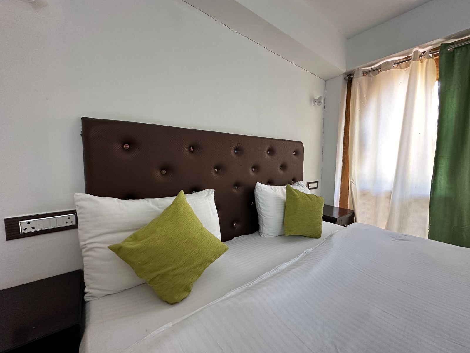 Quarto de hotel com uma cama grande, cabeceira marrom, roupa de cama branca e almofadas verdes.