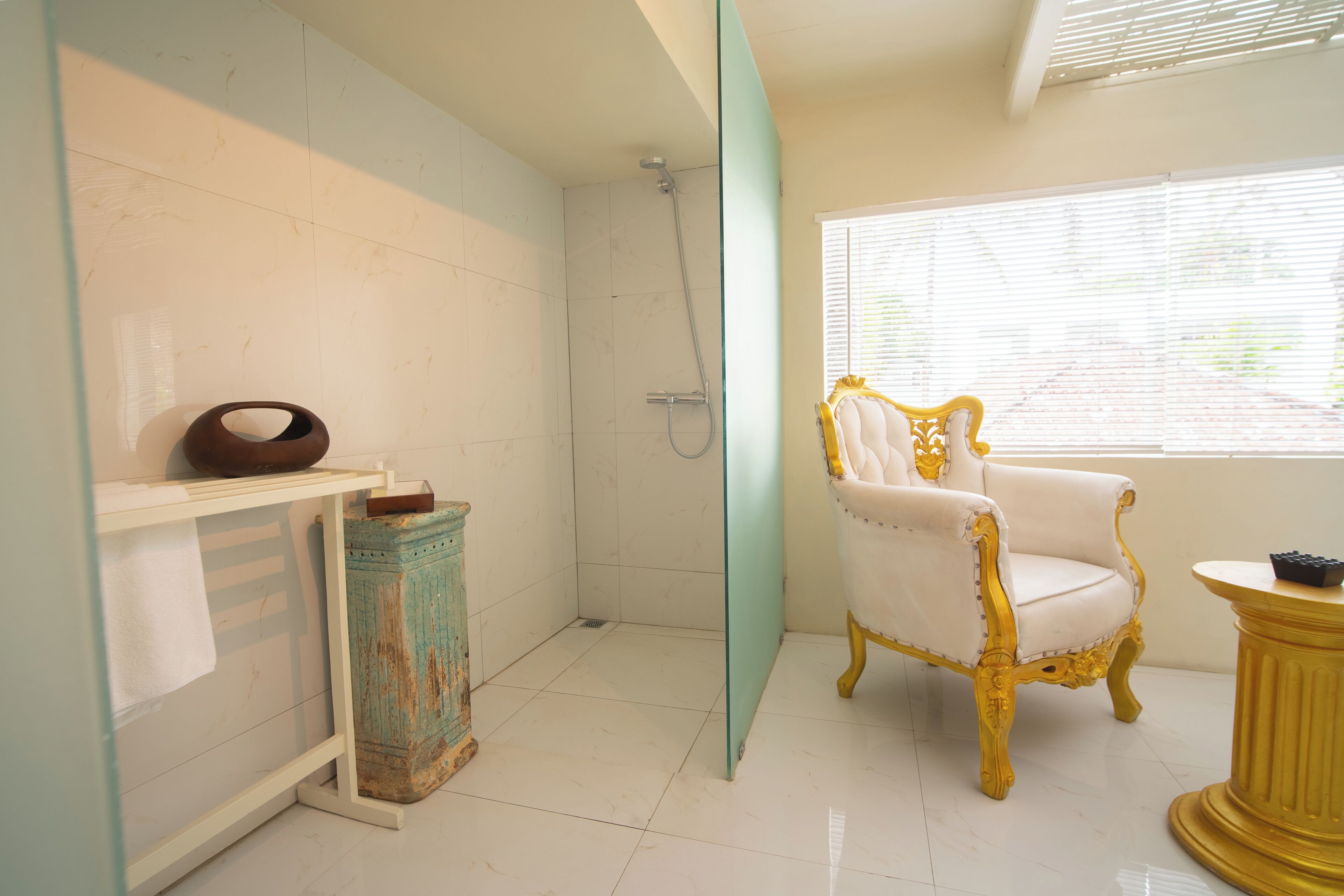 Casa de banho com chuveiro, toucador branco com toalha, cadeira decorativa e janela com estores.