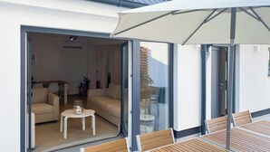 Uma sala de estar moderna com um sofá, uma mesinha e uma porta de vidro que dá para um pátio externo com móveis de madeira e um grande guarda-sol.