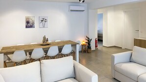 Uma sala de estar moderna com uma mesa de jantar de madeira, cadeiras brancas e um sofá. Há dois aparelhos de ar condicionado fixados na parede e um extintor visível.