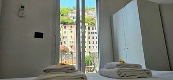 Quarto de hotel com cama, janela com vista para edifícios e termostato fixado na parede.