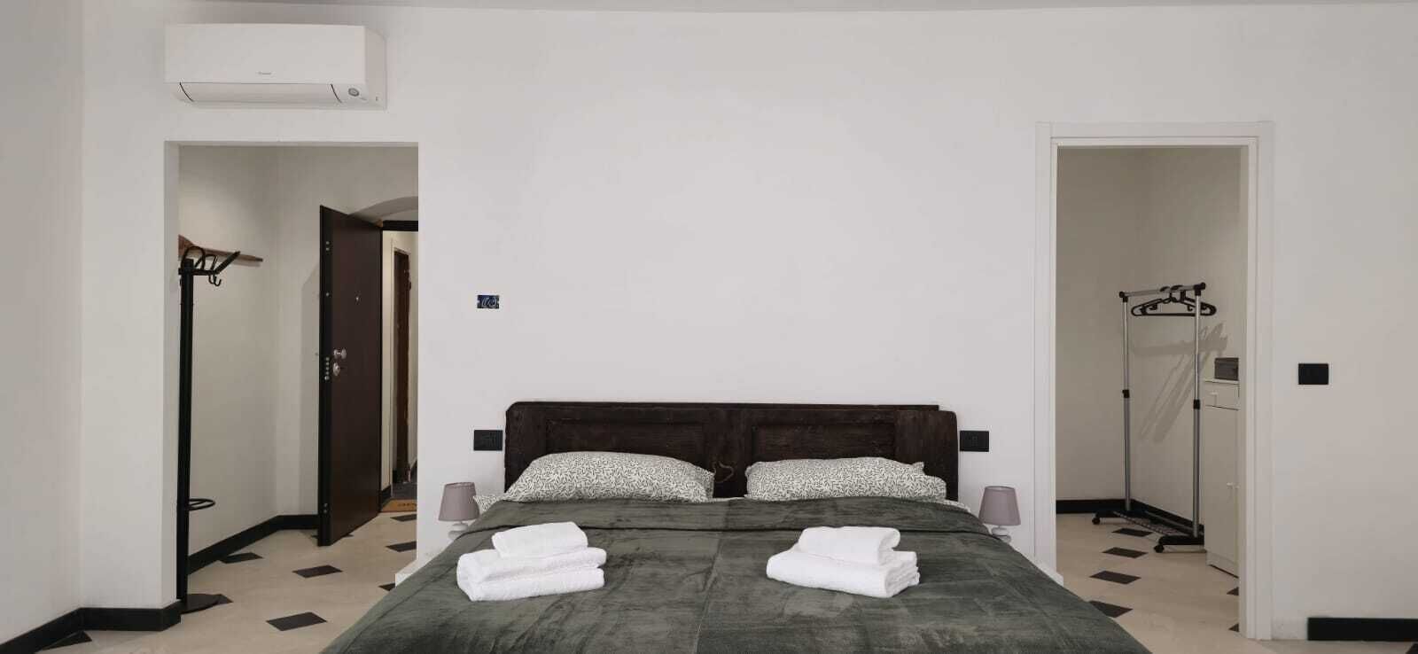 Quarto de hotel com uma cama, duas almofadas, mesa de cabeceira, abajur, cabideiro e porta.