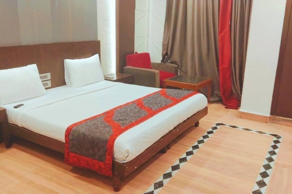Quarto de hotel com uma cama grande, uma mesa de cabeceira com telefone, uma mesinha, uma cadeira e um cobertor vermelho e cinzento.