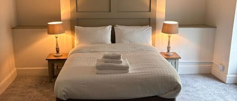 Uma cama bem arrumada com cabeceira, duas mesas de cabeceira com abajures e piso coberto por carpete.