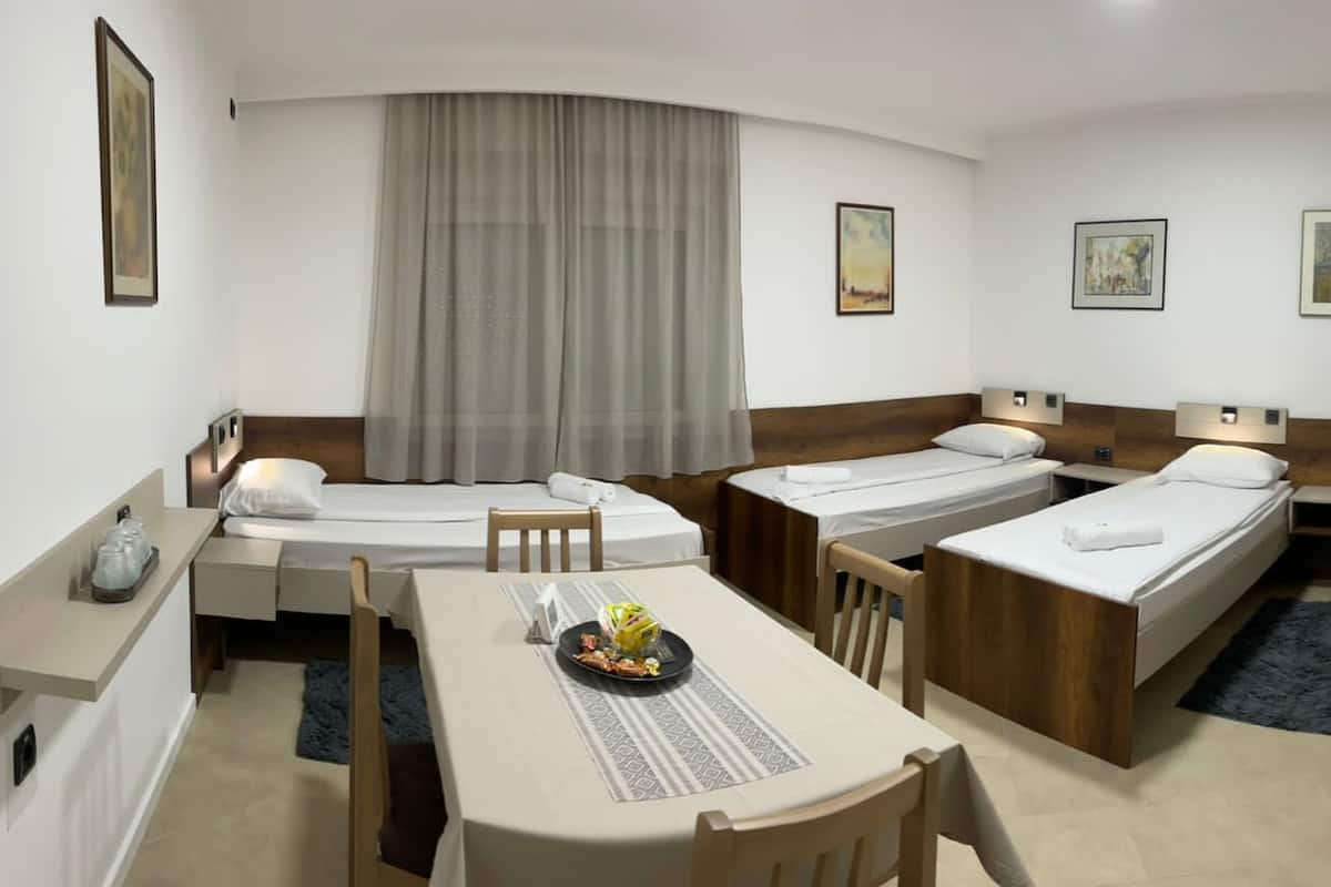 Quarto de hotel com duas camas de solteiro, uma escrivaninha com cadeira, uma mesa de jantar e uma TV fixada na parede.