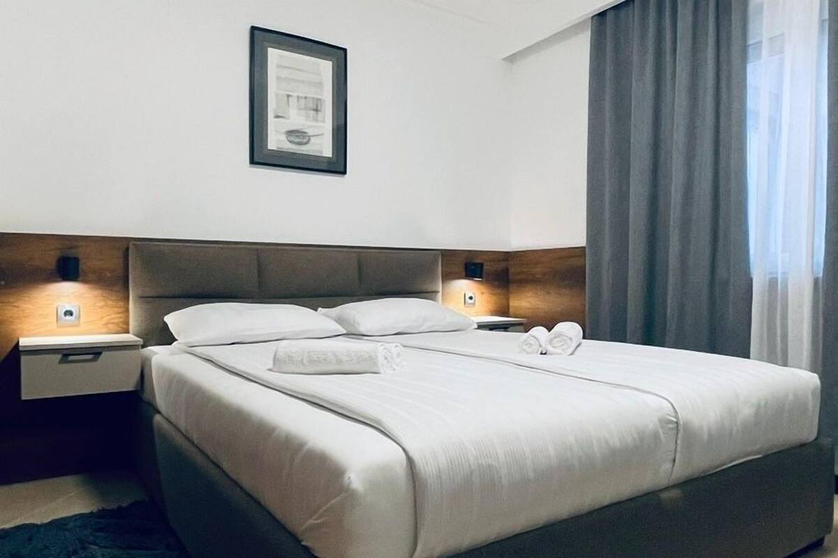 Quarto de hotel moderno com uma cama grande, duas mesas de cabeceira, um quadro na parede e uma janela com cortinas.