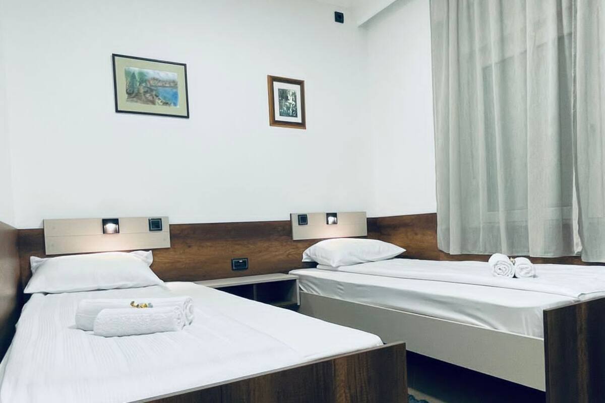 Quarto de hotel com duas camas, uma janela grande com cortinas e duas imagens emolduradas na parede.