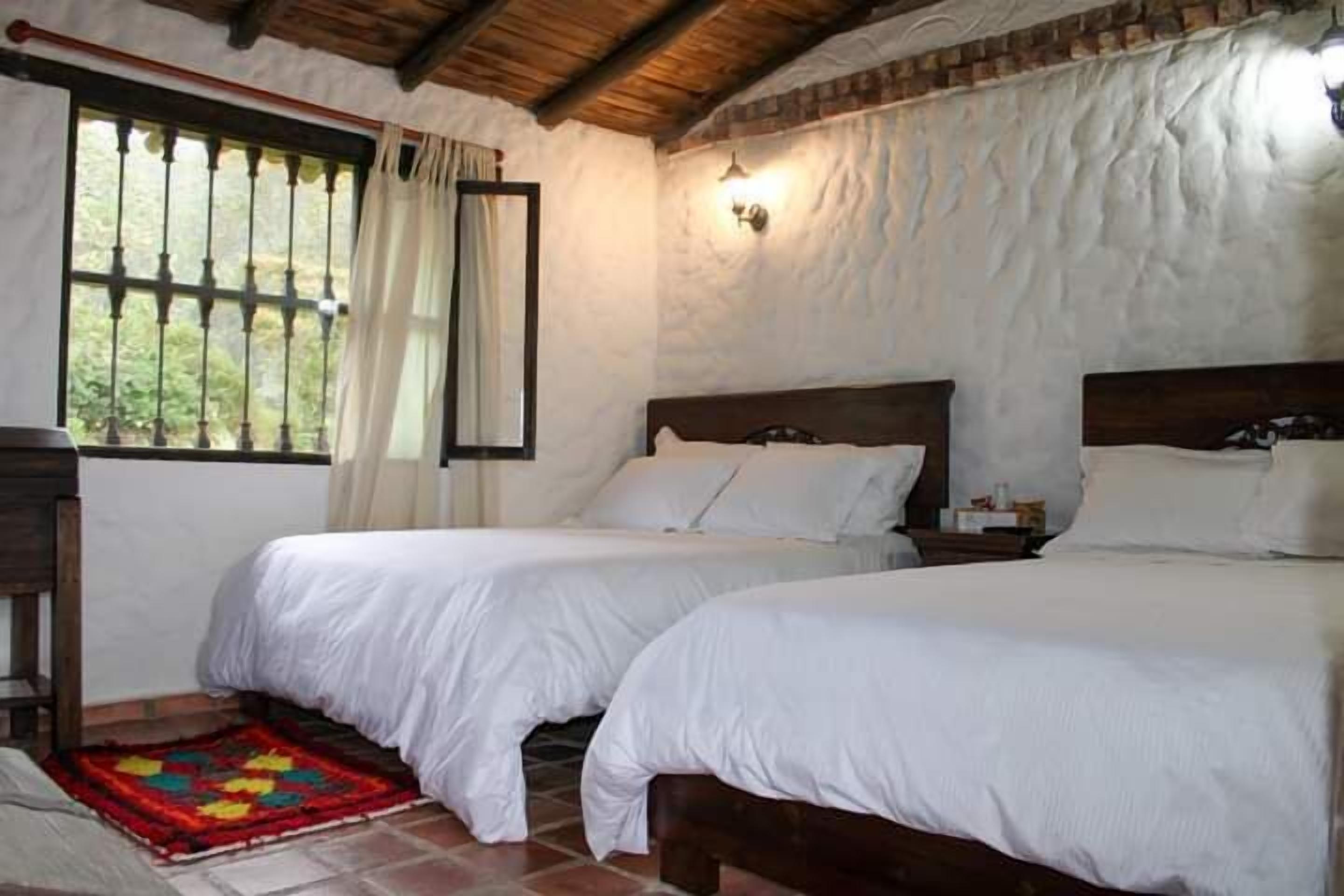 Quarto com duas camas de solteiro, cabeceira de madeira, mesinhas de cabeceira pequenas, janela com grades e um tapete colorido.