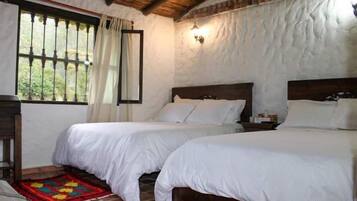 Quarto com duas camas de solteiro, cabeceira de madeira, mesinhas de cabeceira pequenas, janela com grades e um tapete colorido.