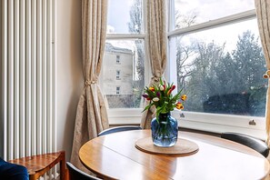 Uma mesa de madeira redonda com um vaso azul contendo tulipas, colocada perto de uma janela com cortinas.