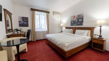 Quarto de hotel com uma cama grande, uma escrivaninha com cadeira, uma mesinha e um abajur.