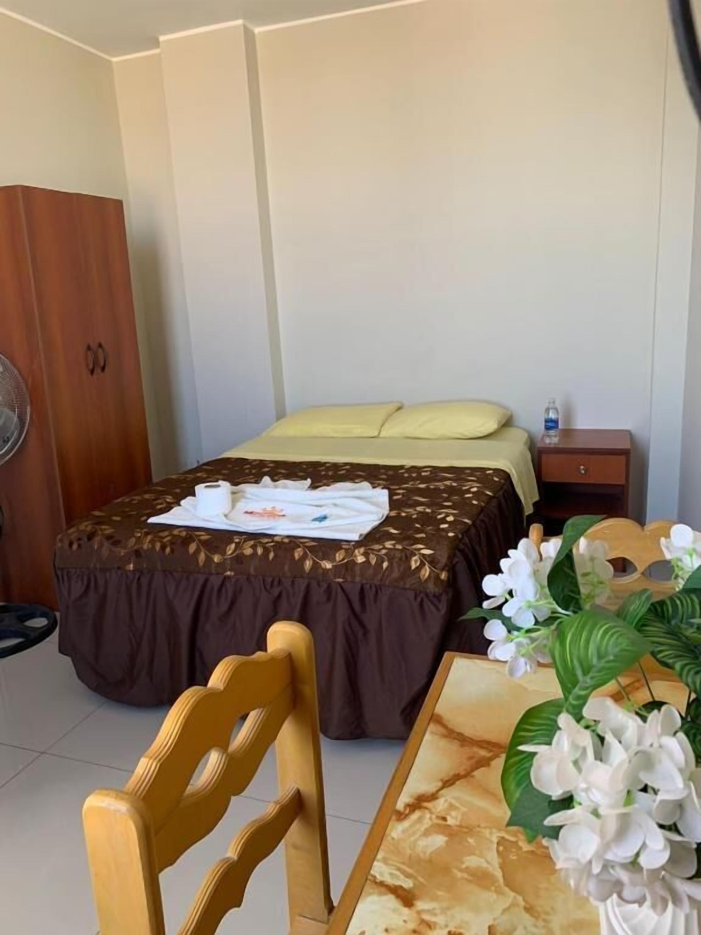 Quarto de hotel com cama, cadeira de madeira, mesa, ventilador e mesa de cabeceira com uma garrafa.