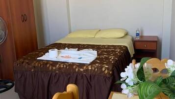 Quarto de hotel com cama, cadeira de madeira, mesa, ventilador e mesa de cabeceira com uma garrafa.