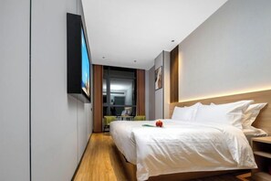 Quarto de hotel moderno com uma cama grande, televisão na parede e um pequeno espaço de estar com mesa e cadeiras.