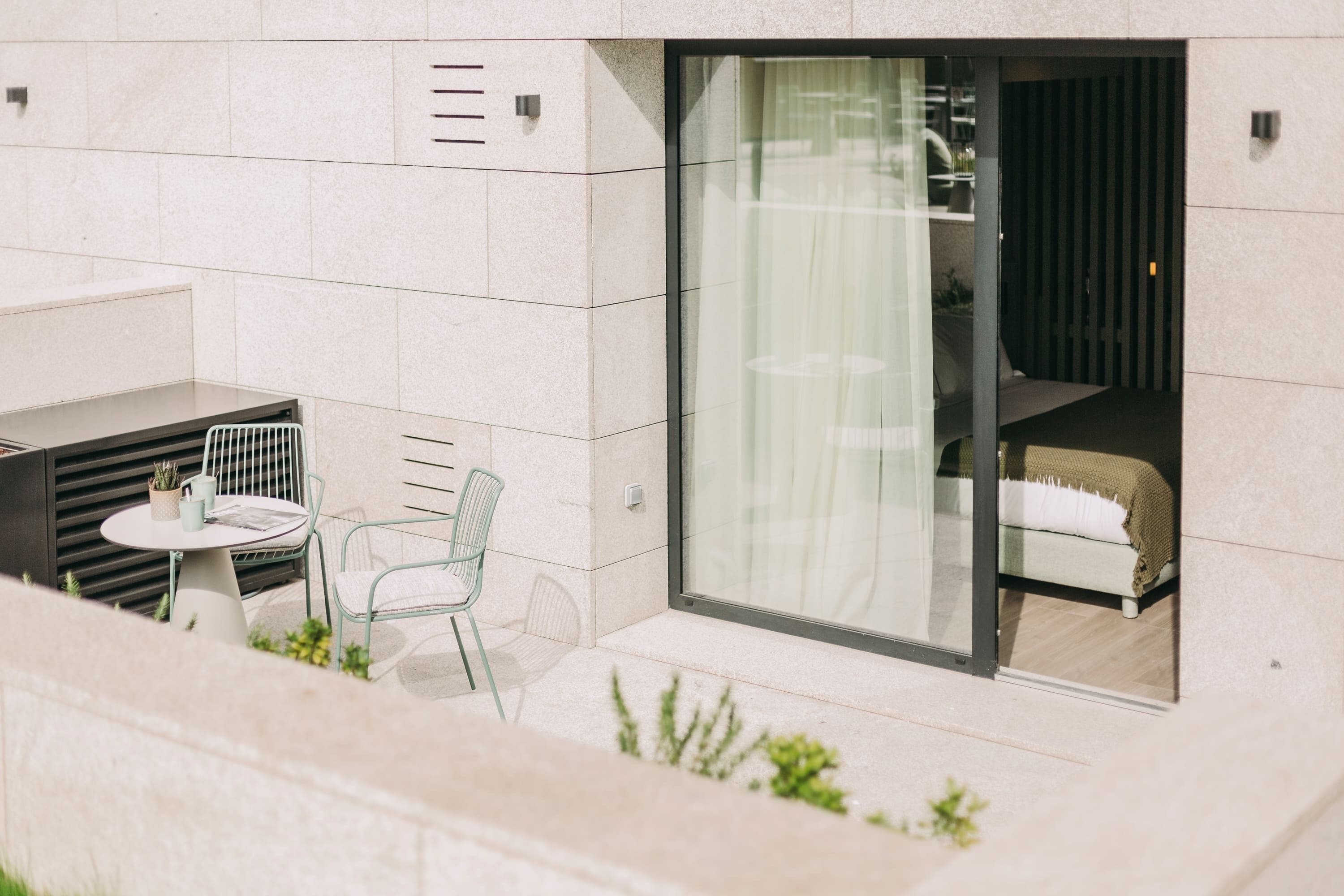 Uma área de estar moderna ao ar livre, com mesa e cadeiras, um pequeno vaso de planta e uma porta de vidro que dá para um quarto com cama e cortinas.