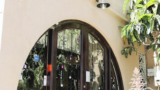 Entrada de um edifício com porta em arco escura, um pequeno jardim com plantas e flores, e um lustre sobre a porta.