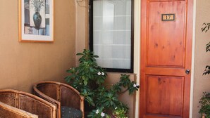 Quarto com porta de madeira numerada 17, cadeiras de vime, mesa de vidro e plantas em vasos.