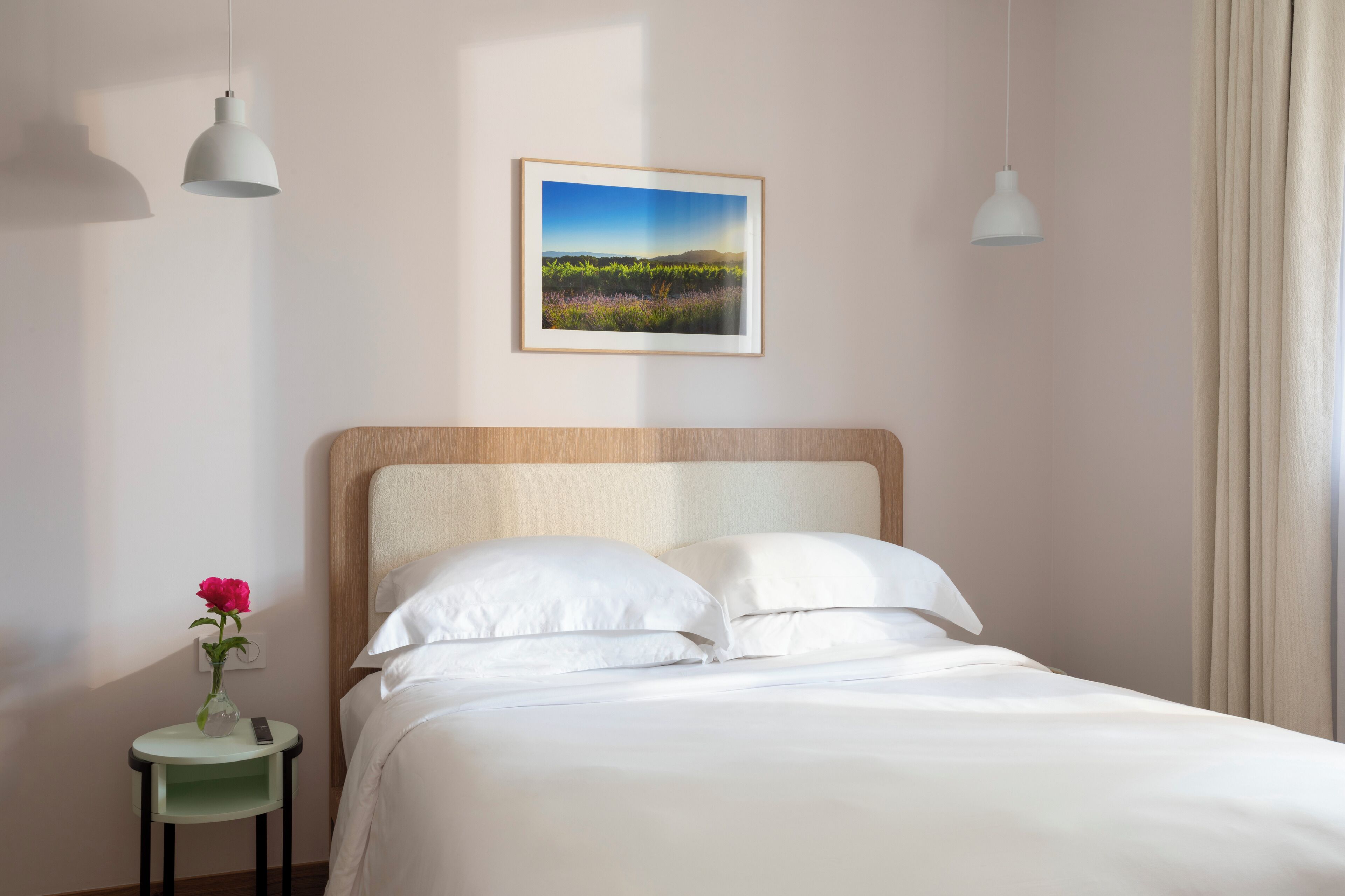 Cama bem arrumada com lençóis brancos, um quadro de paisagem emoldurado acima e uma pequena mesa com um vaso de flores ao lado da cama.