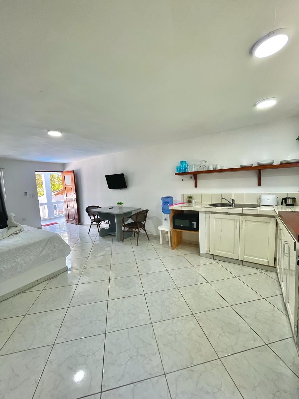 Quarto de hotel com cama, mesa de jantar, cadeiras, cozinha com armários e pia, e televisão na parede.