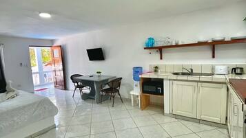 Quarto de hotel com cama, mesa de jantar, cadeiras, cozinha com armários e pia, e televisão na parede.