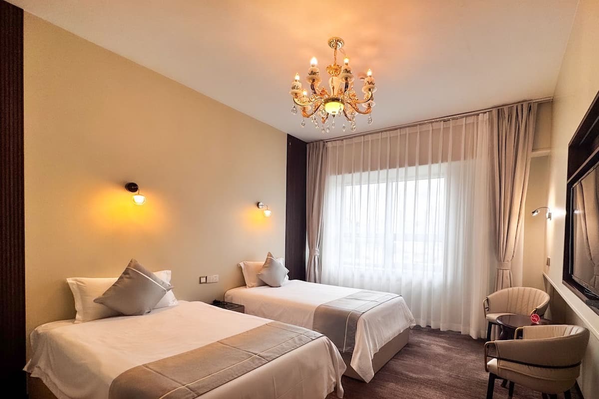 Quarto de hotel com duas camas, um candelabro, uma janela grande com cortinas e uma cadeira.